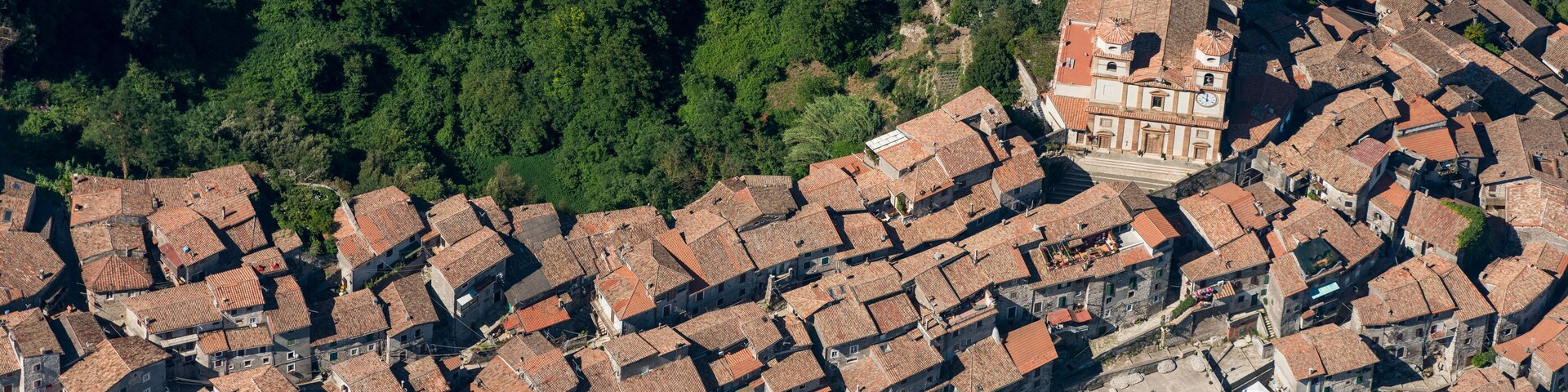 Vista aérea de uma aldeia densamente povoada com edifícios de telhados em terracota, cercada por uma floresta verde exuberante.