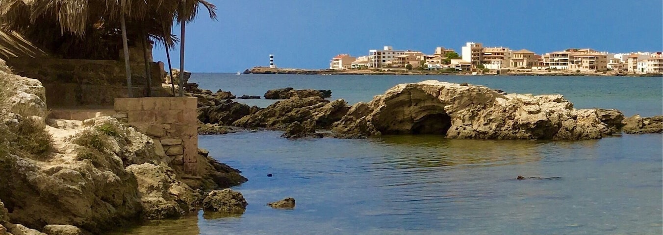 Uma praia com águas cristalinas, um afloramento rochoso e um horizonte urbano distante.