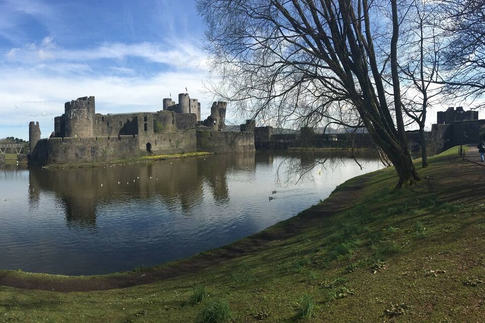 Um castelo histórico junto a um lago tranquilo, com um primeiro plano gramado e árvores sem folhas.