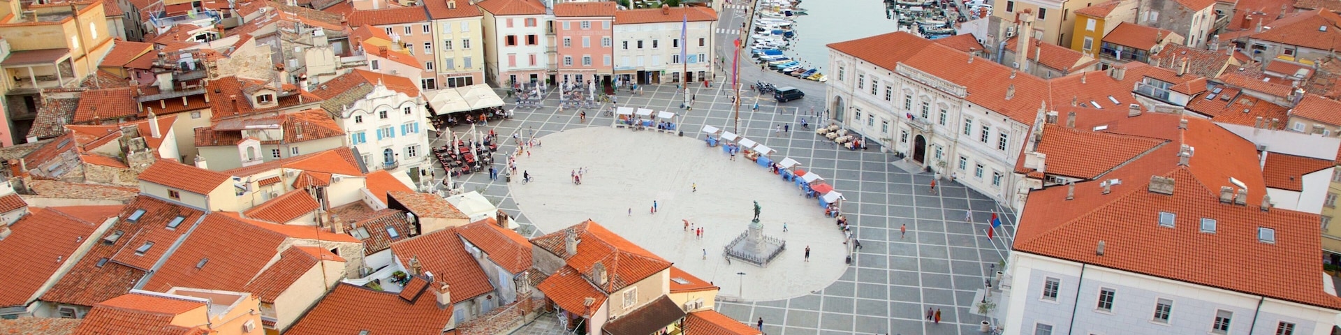 Uma vila costeira com telhados vermelhos, uma praça central e uma marina com barcos.