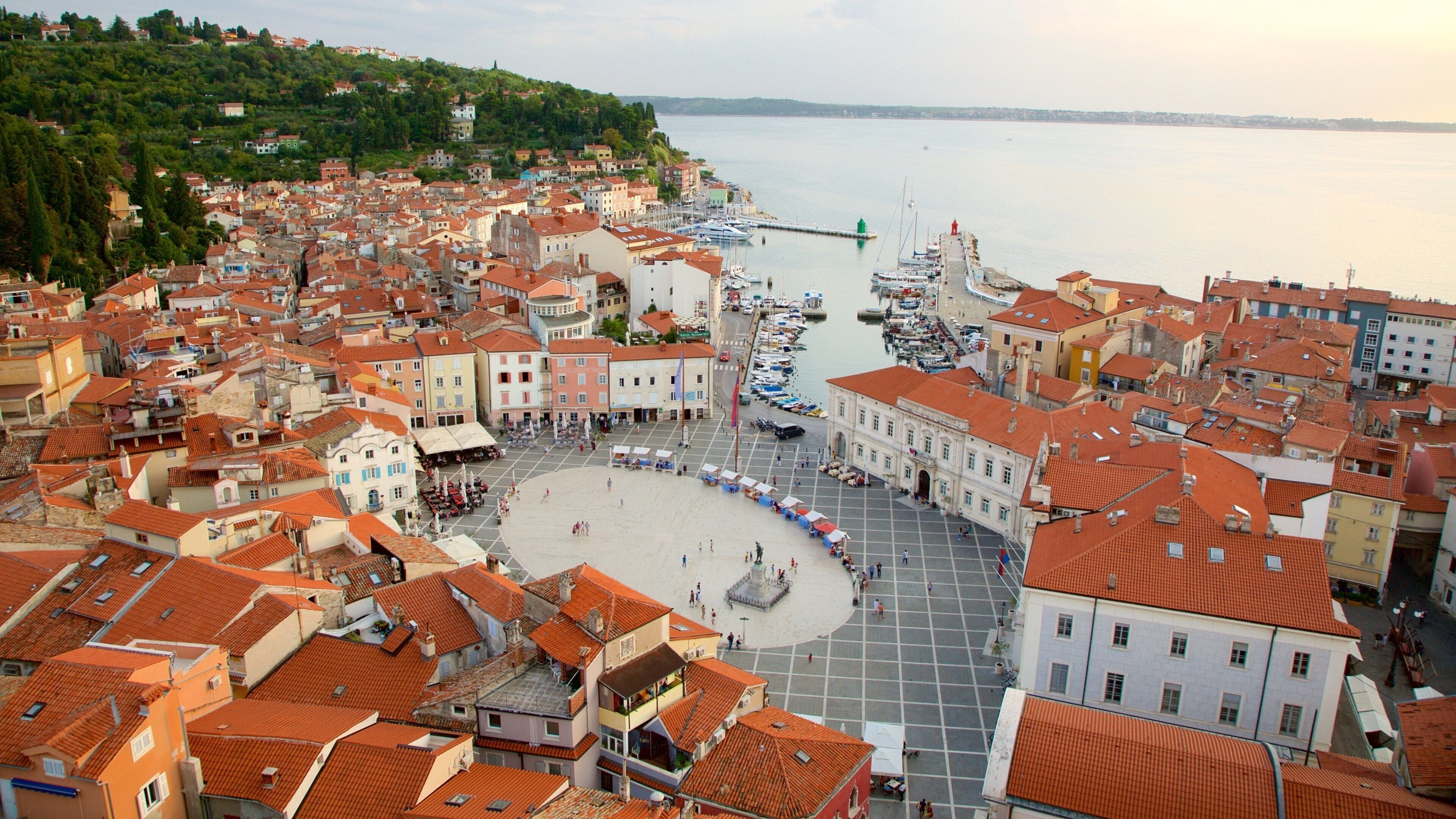 Uma cidade costeira com edifícios de telhado vermelho, uma praça central e um porto com barcos.