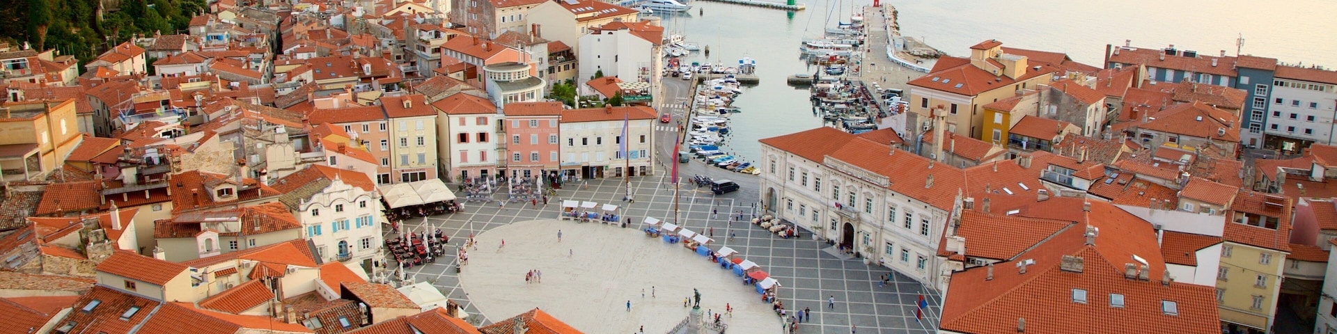 Uma cidade costeira com edifícios de telhado vermelho, uma praça central e um porto com barcos.