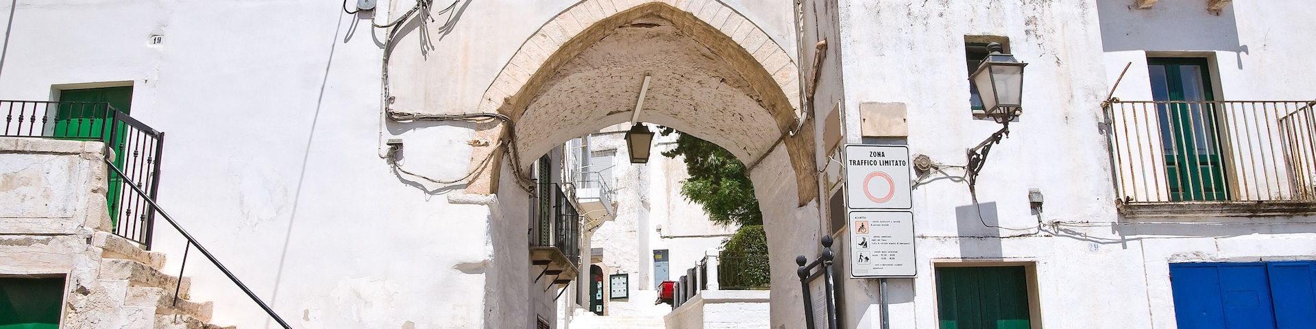Uma rua estreita com um arco de pedra, edifícios brancos com persianas verdes e uma porta azul.
