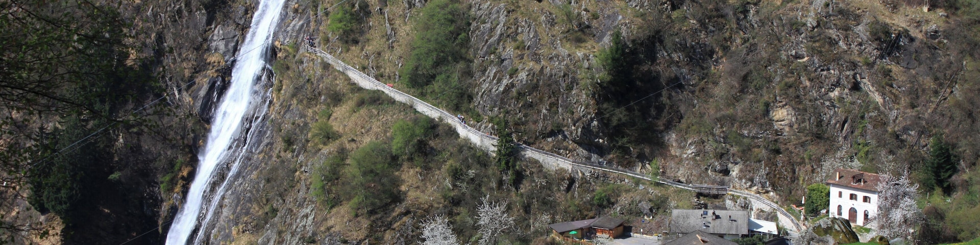 Uma cascata desce por um declive rochoso, uma estrada sinuosa e um conjunto de edifícios com uma estrutura branca destacada.