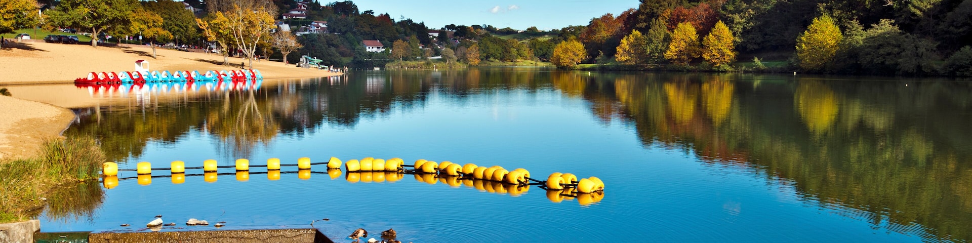 Um lago tranquilo com um píer, coloridos barcos a remo e árvores de outono refletidas na água.