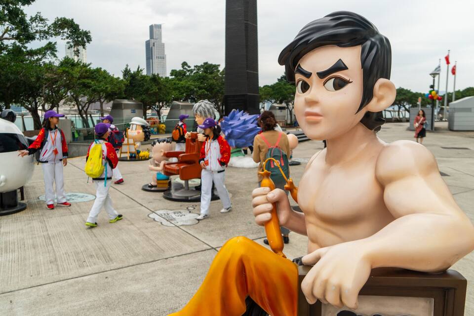 Estátua de uma figura musculosa segurando um bastão, cercada por crianças e adultos em uma praça pública com árvores e edifícios ao fundo.