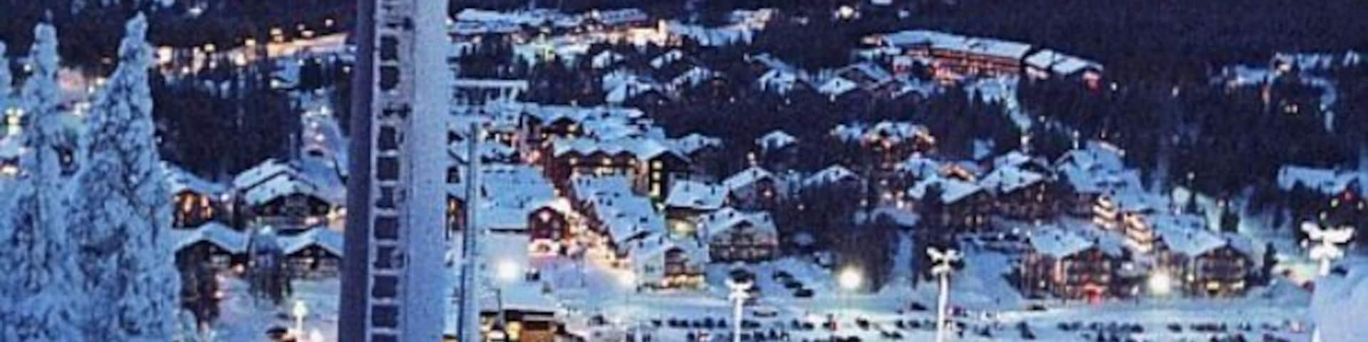 Estância de esqui coberta de neve ao entardecer, com edifícios iluminados e um teleférico.