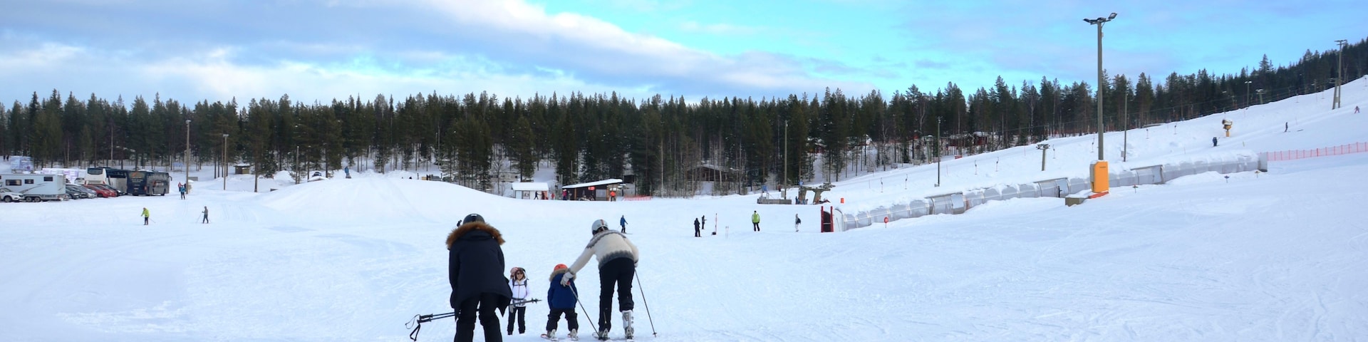 Pista de esqui coberta de neve, com esquiadores e uma área arborizada ao fundo.