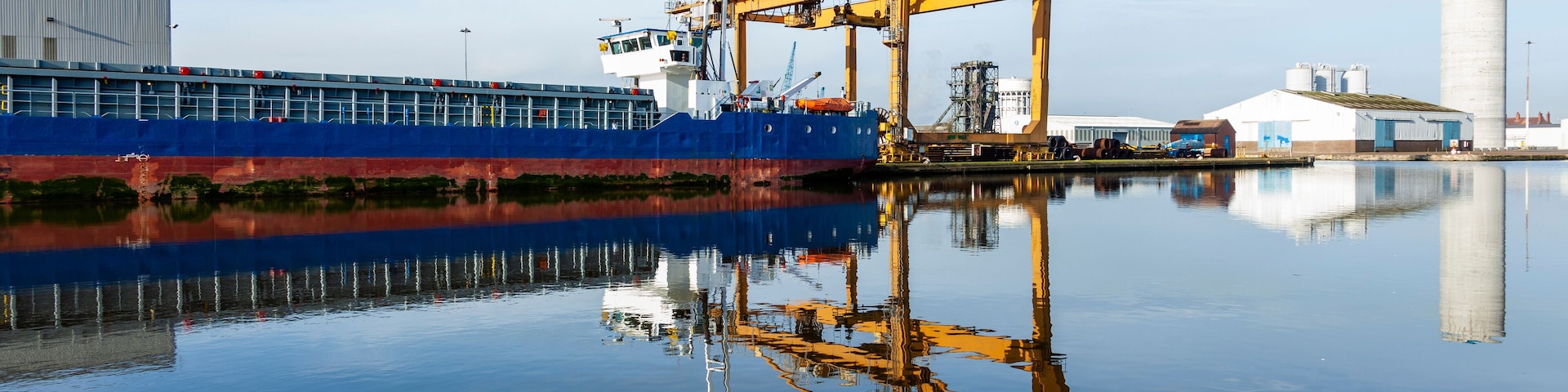 Um navio cargueiro atracado em um porto, com guindastes e edifícios industriais ao fundo.