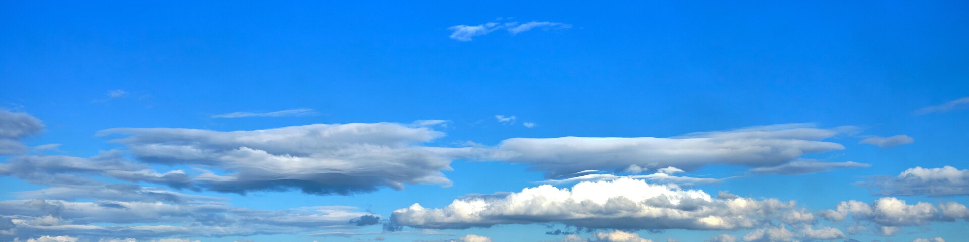 Céu azul límpido com nuvens dispersas, uma lua distante e uma vasta paisagem com colinas ondulantes e um corpo d'água.