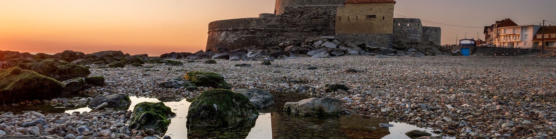 Uma fortaleza de pedra histórica com telhado vermelho, rodeada por pedregulhos e uma poça de água límpida que reflete a estrutura.