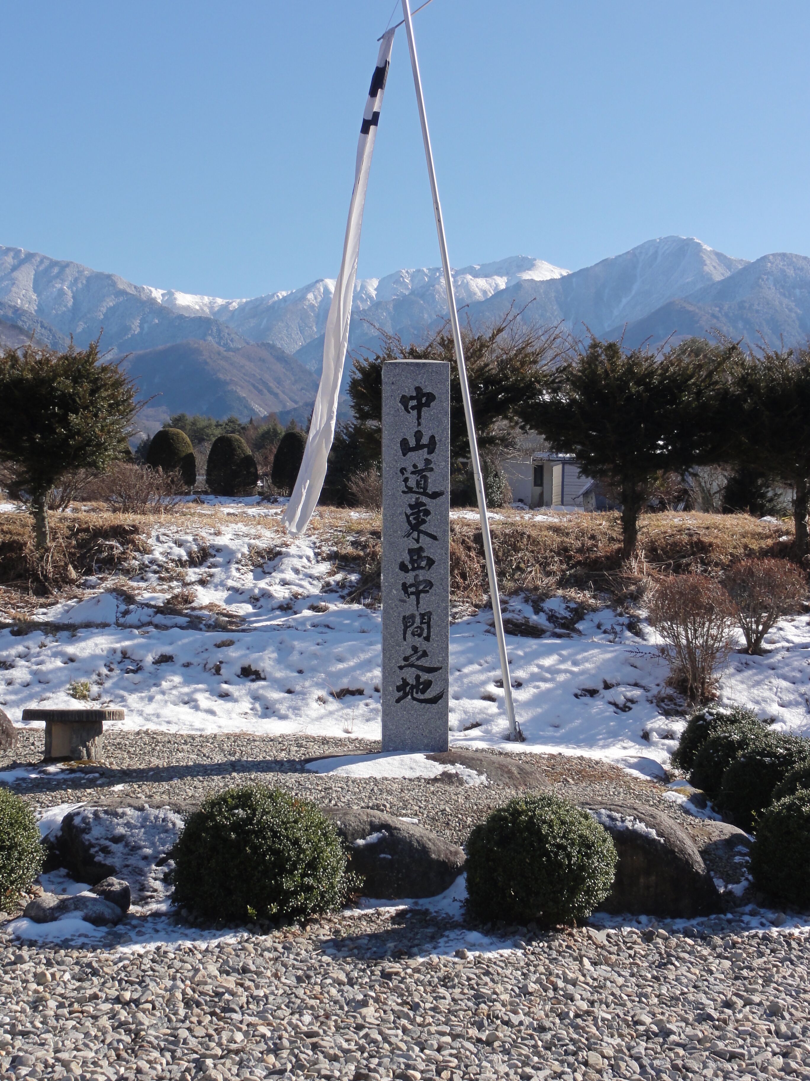 Um monumento de pedra com caracteres japoneses, rodeado por terreno coberto de neve, rochas e árvores, com montanhas ao fundo.