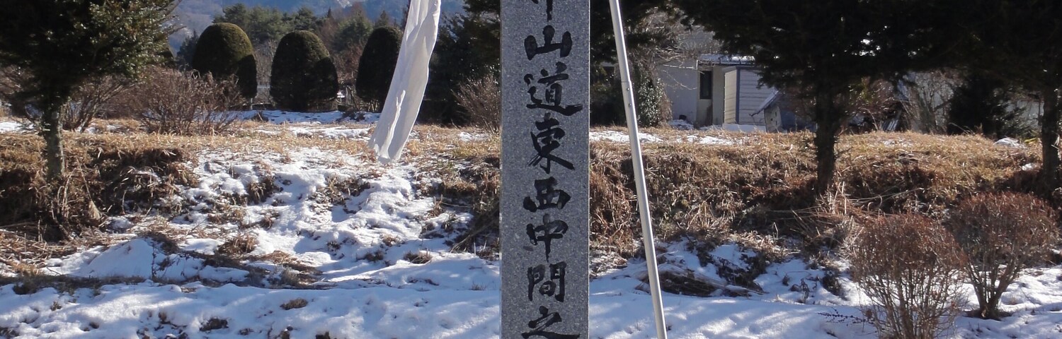 Um monumento de pedra com caracteres japoneses, rodeado por terreno coberto de neve, rochas e árvores, com montanhas ao fundo.