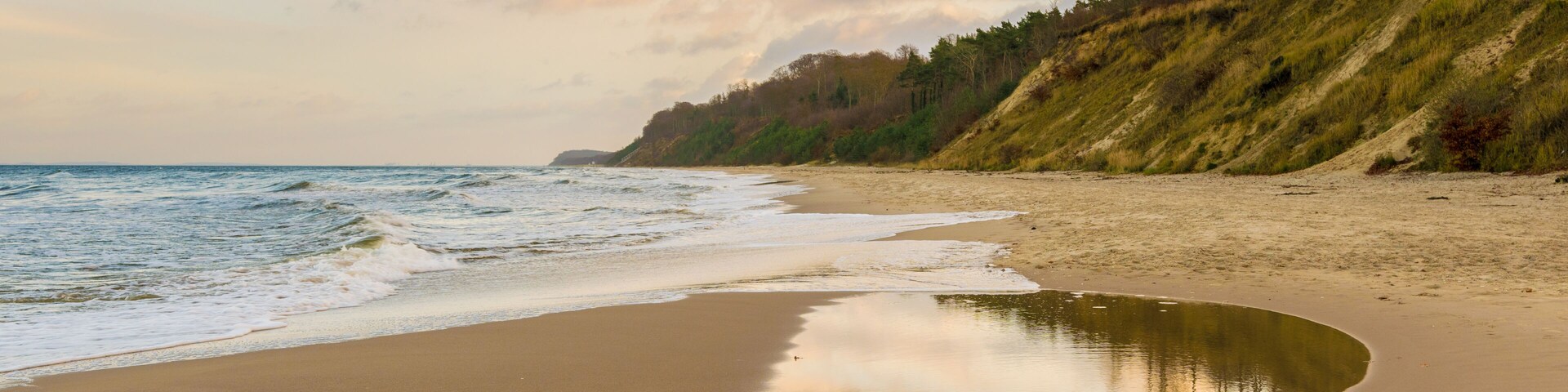 Uma praia arenosa com ondas suaves, céu claro e um morro coberto de árvores.