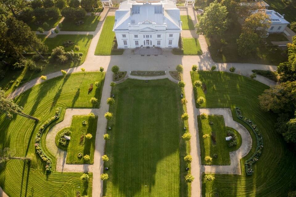 Vista aérea de uma grande propriedade simétrica, com gramados bem cuidados, um jardim circular central e um corpo d'água ao fundo.