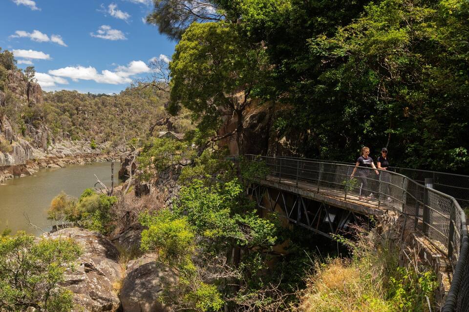Uma ponte suspensa sobre um rio, rodeada por penhascos rochosos e vegetação densa.