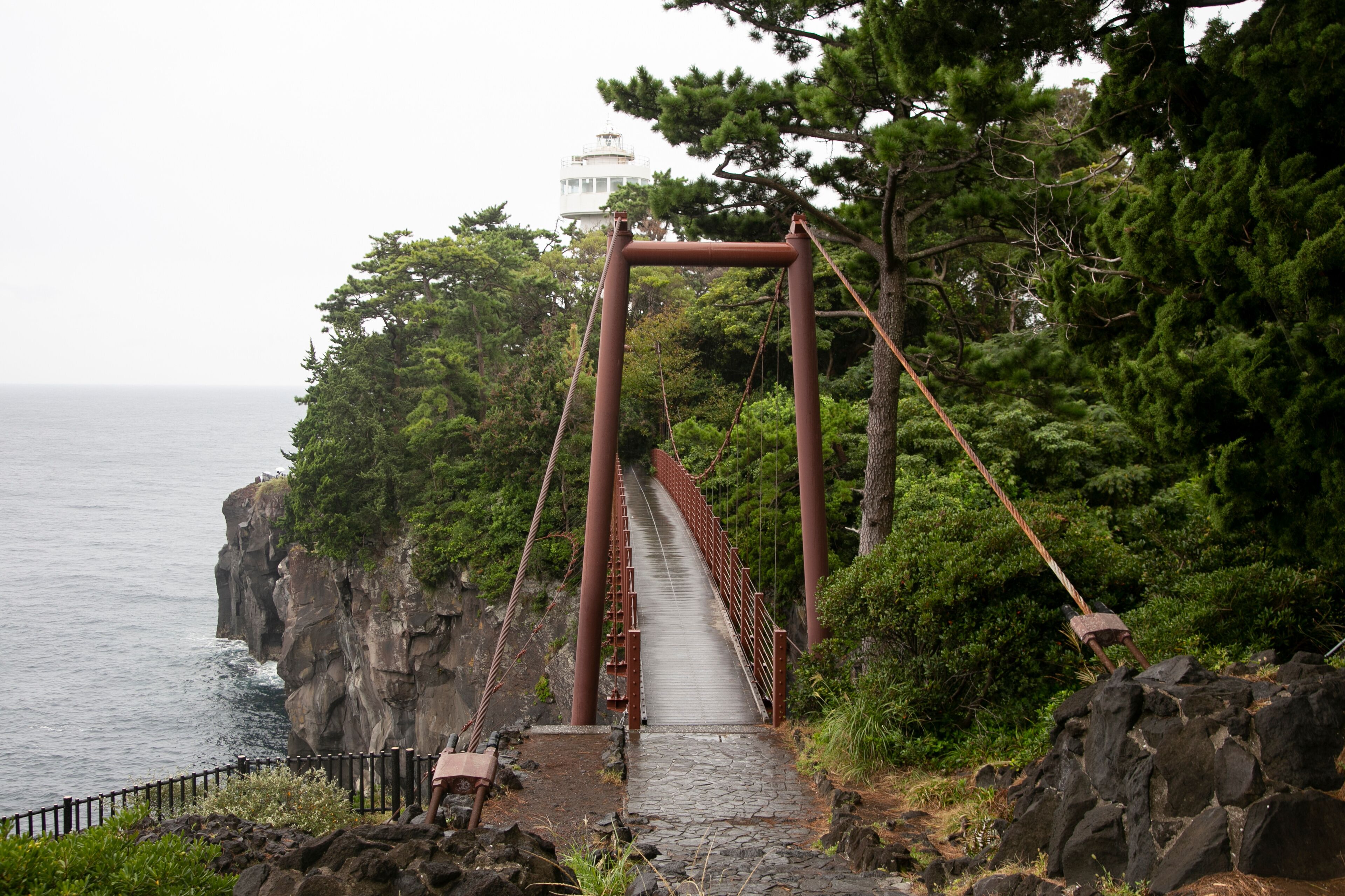 Uma ponte suspensa sobre um penhasco rochoso, com um farol ao fundo.