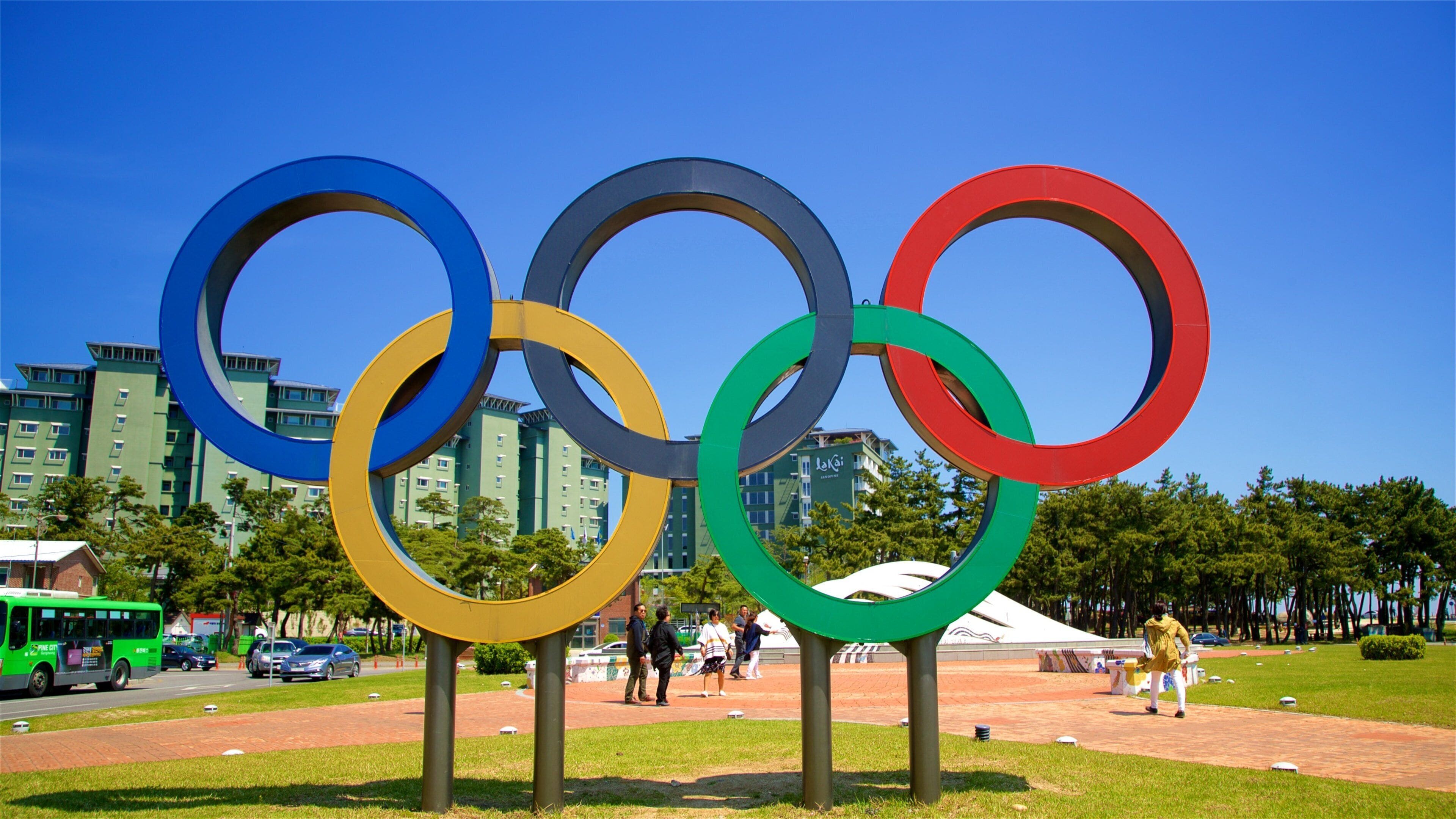 Escultura dos anéis olímpicos em um parque, com edifícios e árvores ao fundo.