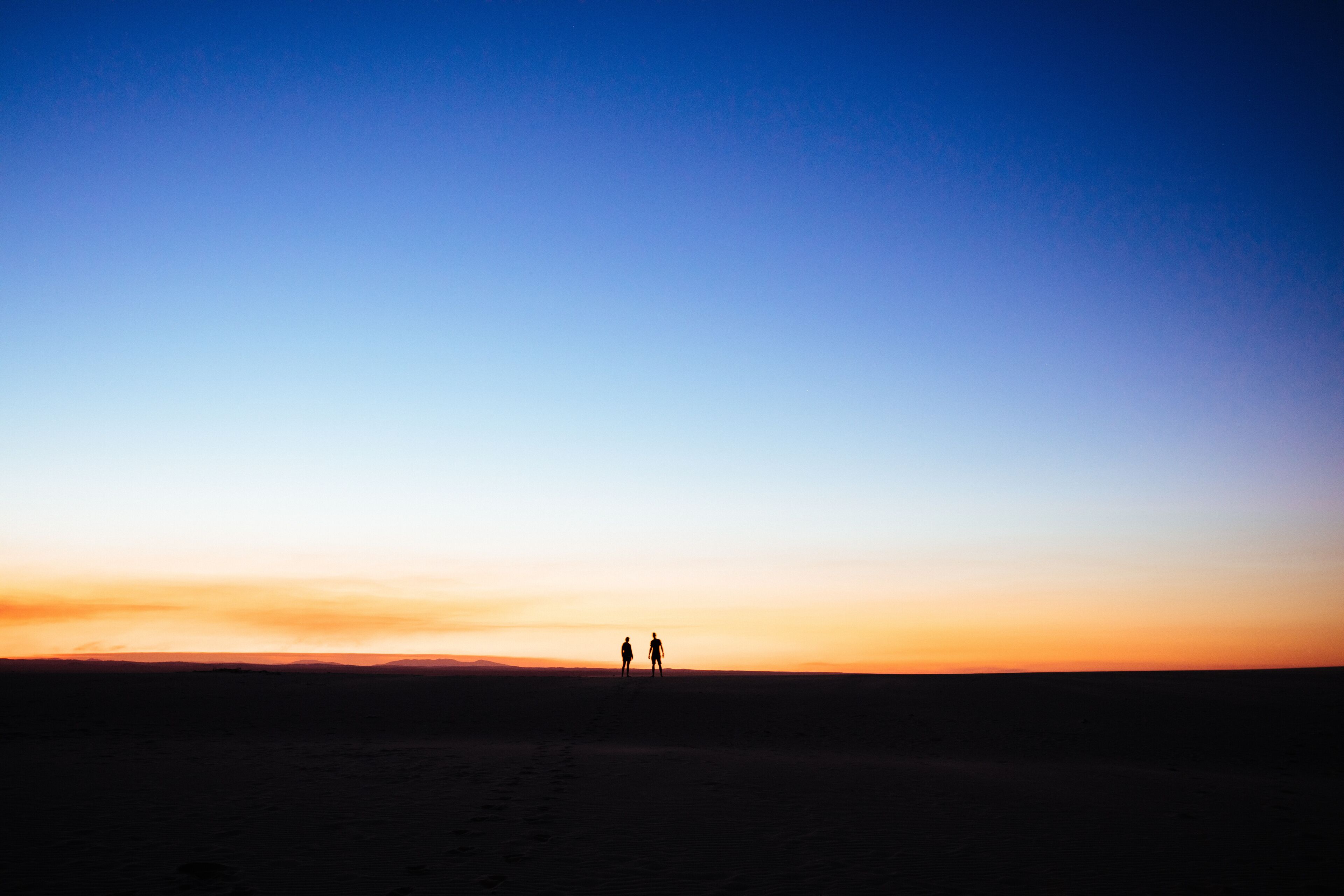 Paisagem desértica ao pôr do sol, com duas silhuetas de pessoas caminhando.