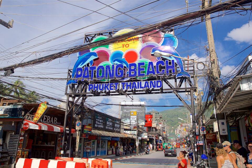 Rua movimentada com uma placa colorida que diz "Bangkok Beach Phuket Thailand" e diversos estabelecimentos e veículos.