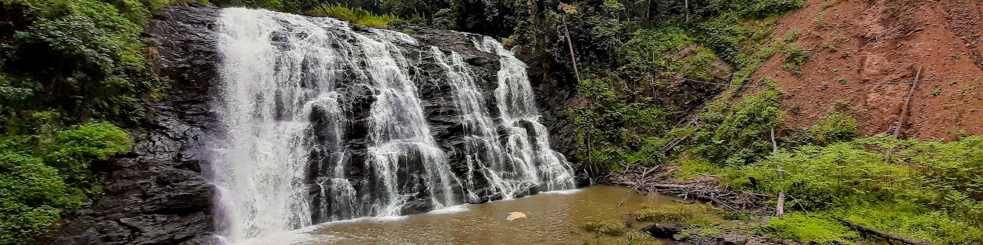 Uma cascata despenca num rio, rodeada por vegetação exuberante e uma encosta vermelha.