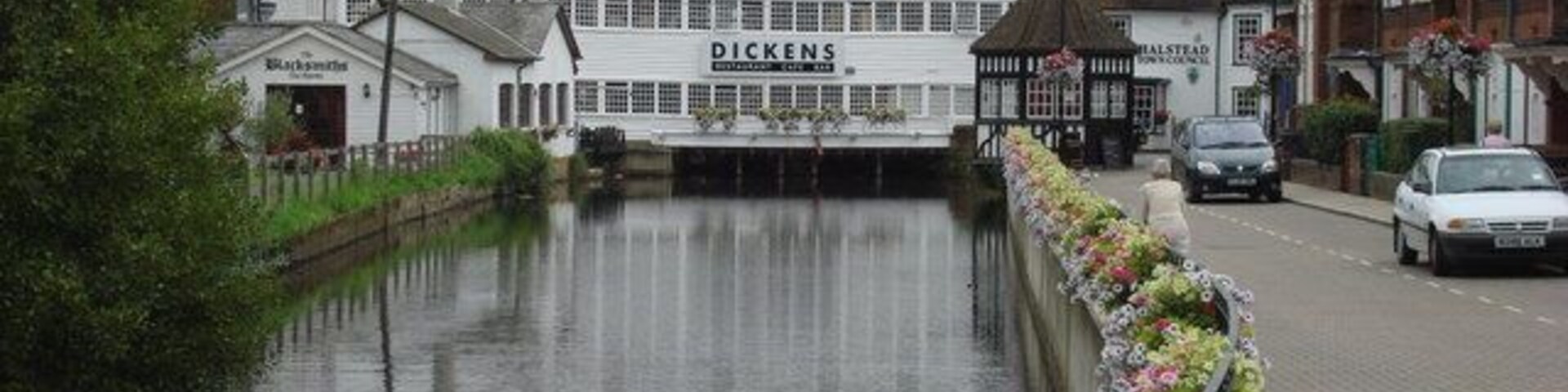 Um canal com um pato nadando, um prédio branco com a inscrição 'DICKENS' e uma fileira de flores ao longo da margem do canal.