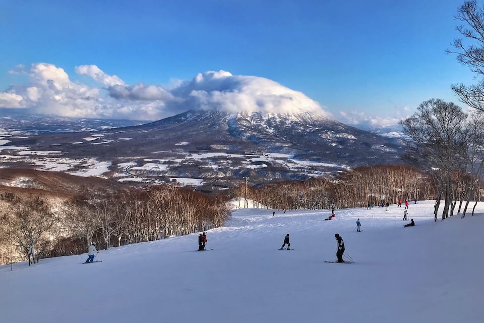 Pista de esqui coberta de neve, com esquiadores, uma montanha ao fundo e céu azul límpido.