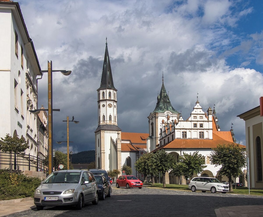 Uma igreja histórica com uma torre do relógio alta e uma agulha menor, rodeada por ruas de calçada e carros estacionados.