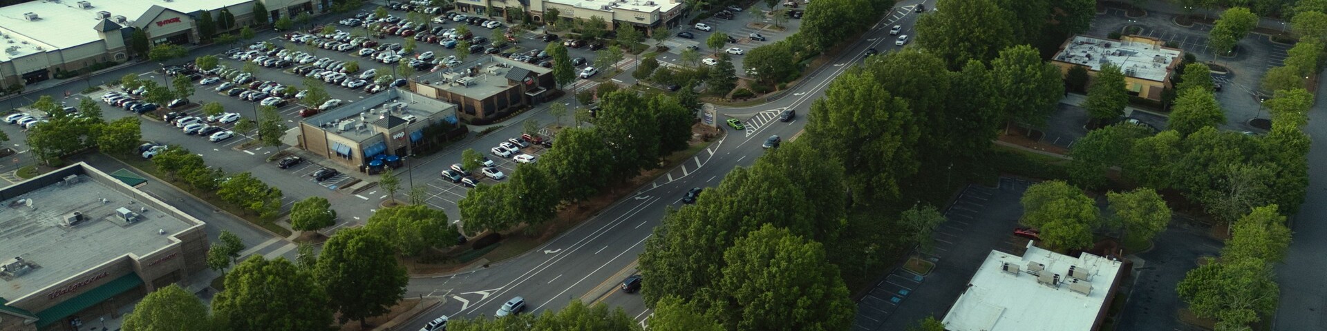 Vista aérea de uma área comercial com vários estacionamentos, uma via principal e vegetação ao redor.