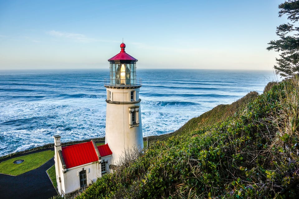 Um farol com teto vermelho, um edifício branco com teto vermelho e o oceano ao fundo.