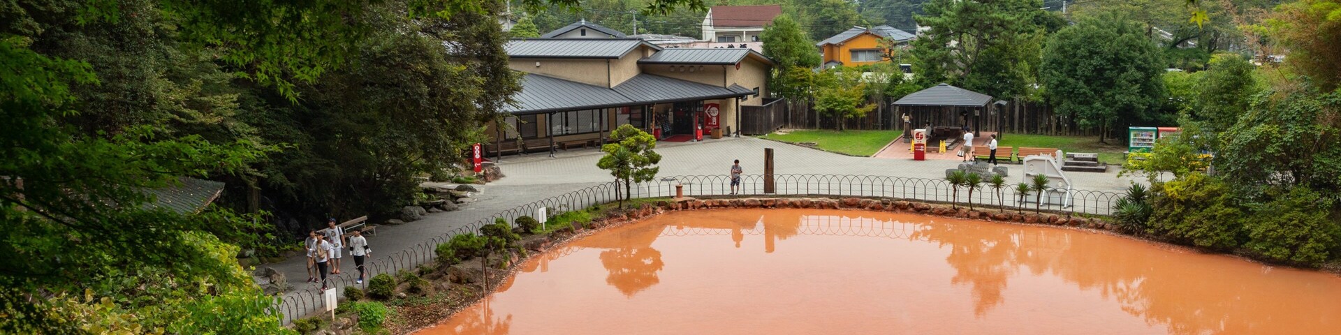 Um lago com tonalidade avermelhada, rodeado por vegetação exuberante e alguns edifícios ao fundo.