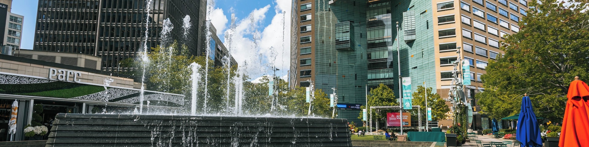 Uma praça urbana com fonte, edifícios modernos e mesas ao ar livre.