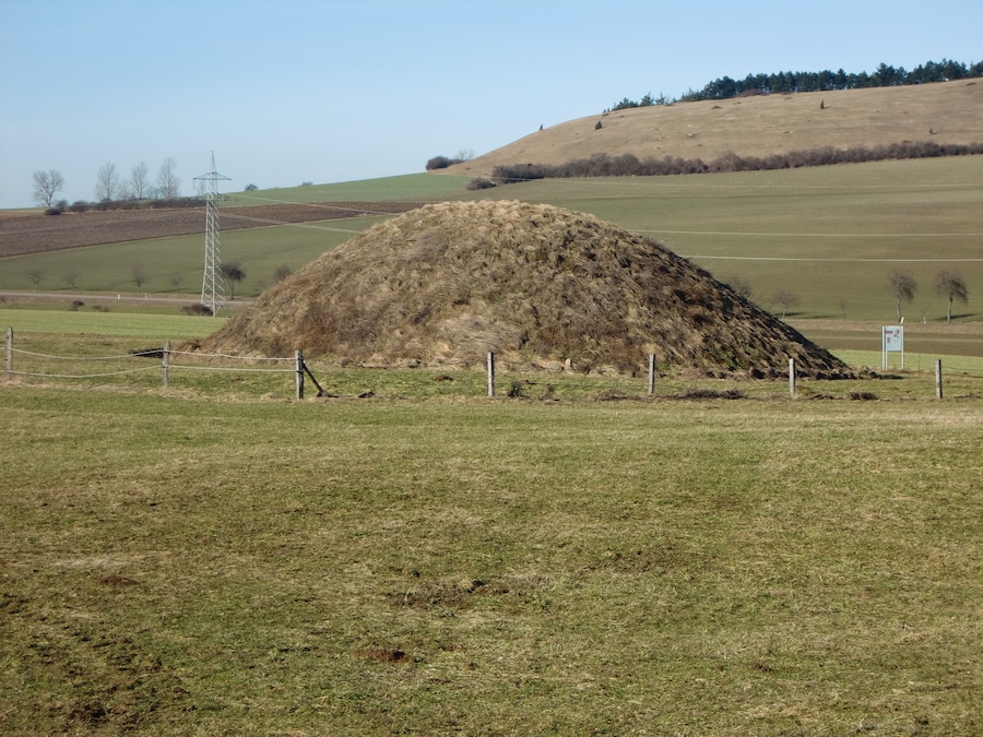 Uma grande pilha de feno em um campo, com uma cerca e uma torre de linhas de energia ao fundo.