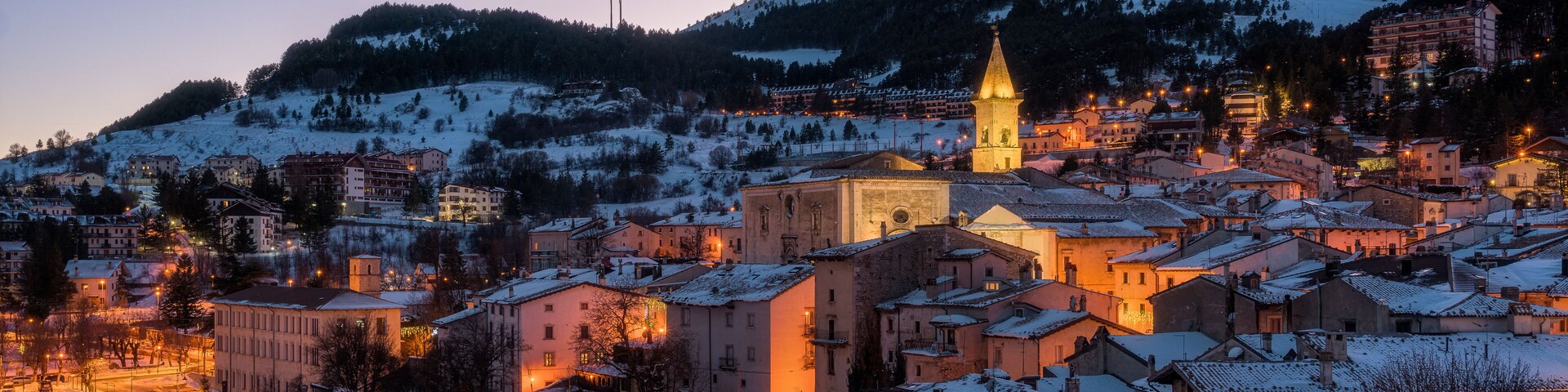 Uma cidade coberta de neve ao entardecer, com edifícios iluminados e uma torre de igreja imponente.