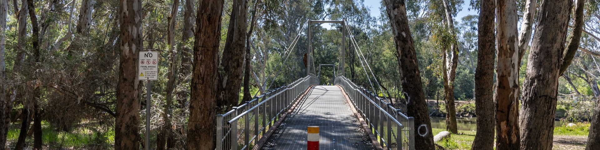 Uma ponte para pedestres numa floresta, com árvores e uma placa de 'Proibido o Acesso'.