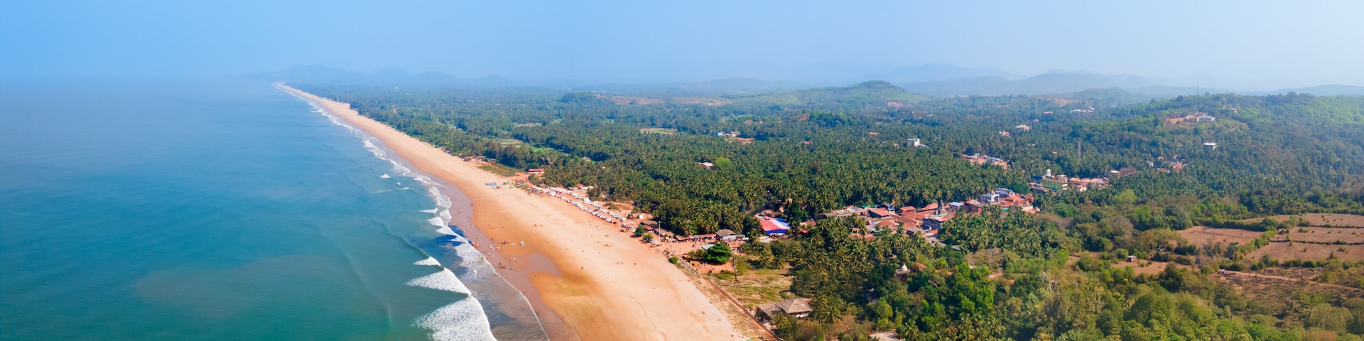 Paisagem costeira com uma praia de areia, céu azul límpido e colinas verdes ao fundo, com alguns edifícios espalhados.