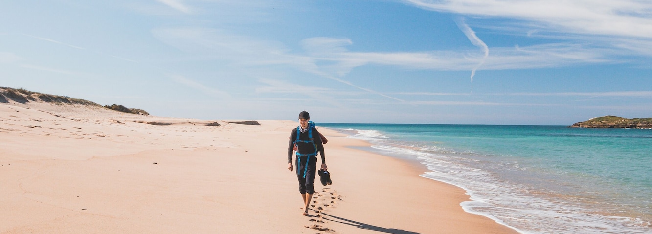 Uma pessoa caminhando na praia arenosa, com pegadas que se estendem em direção ao mar.