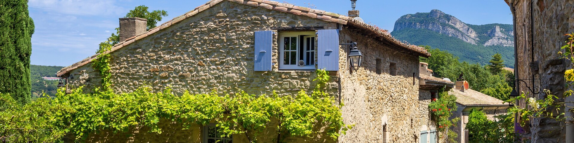 Uma casa de pedra com telhado de colmo, rodeada por vegetação exuberante e flores vermelhas.