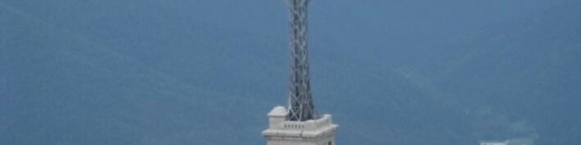 Um monumento em forma de cruz numa colina, com o mar ao fundo.