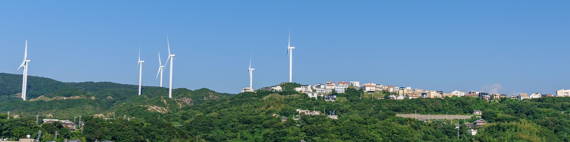 Uma cidade costeira com turbinas eólicas numa colina e um cais que se estende para a água.