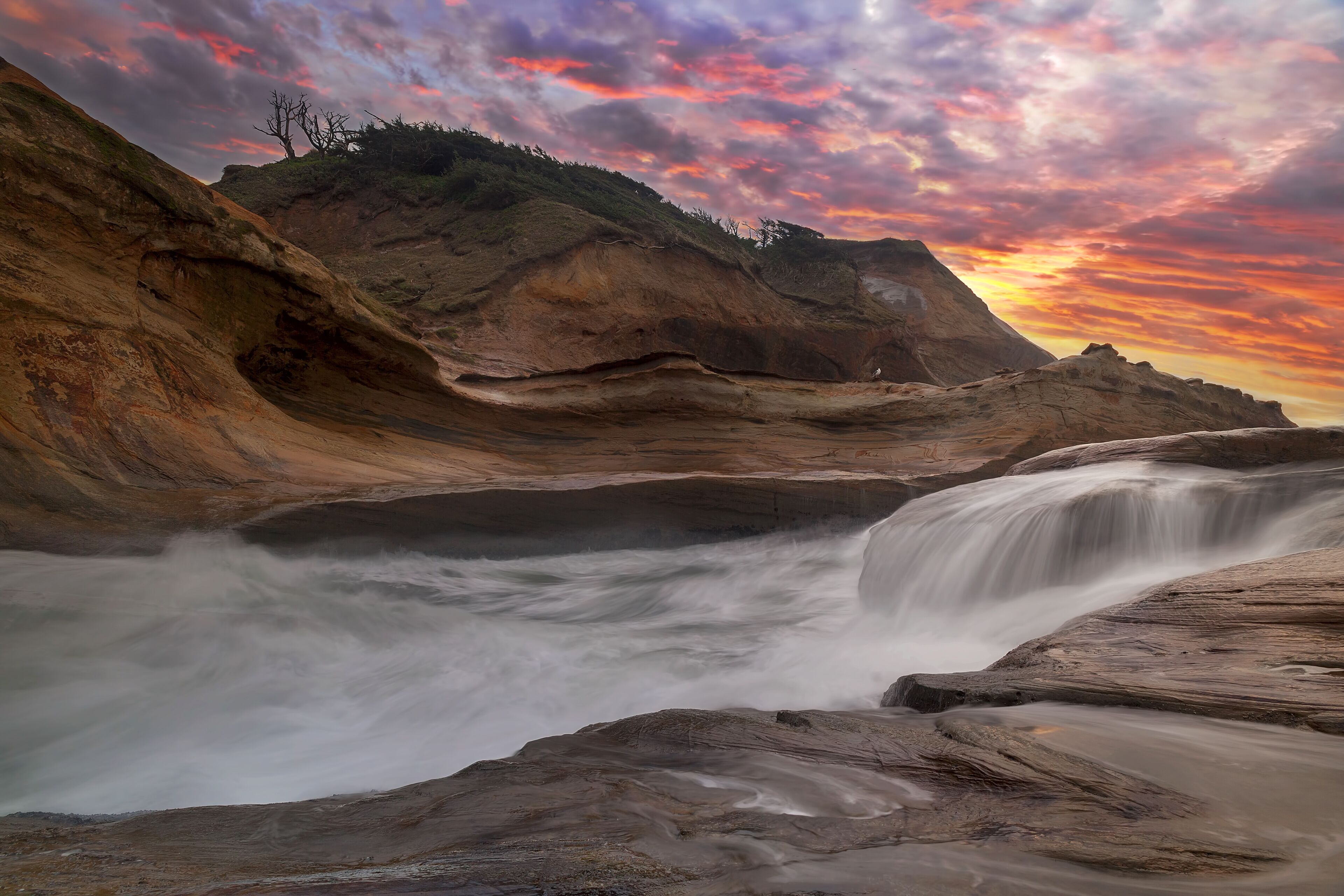 Uma cascata desce sobre um terreno rochoso, com um pôr do sol ao fundo.
