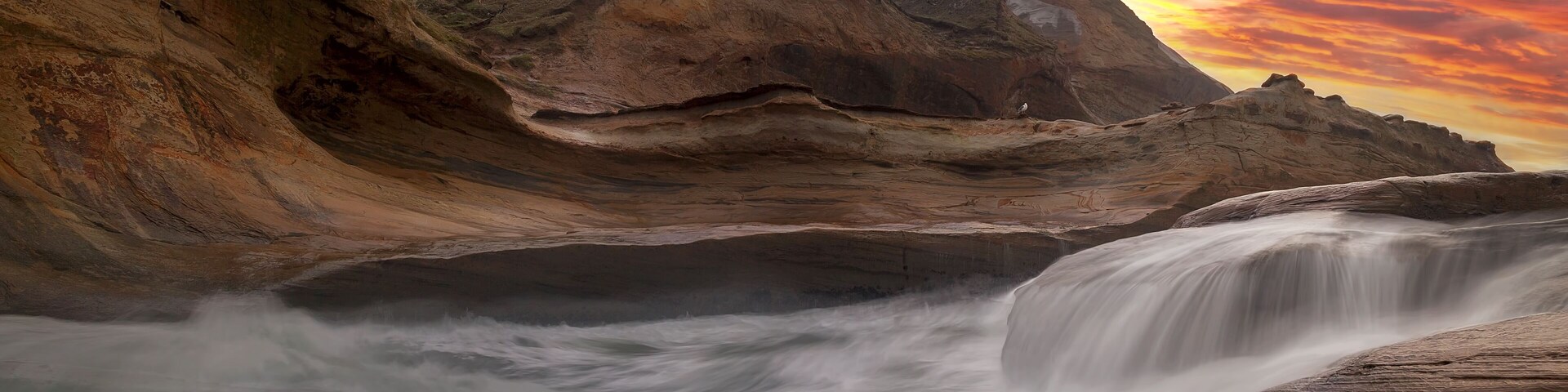 Uma cascata desce sobre um terreno rochoso, com um pôr do sol ao fundo.