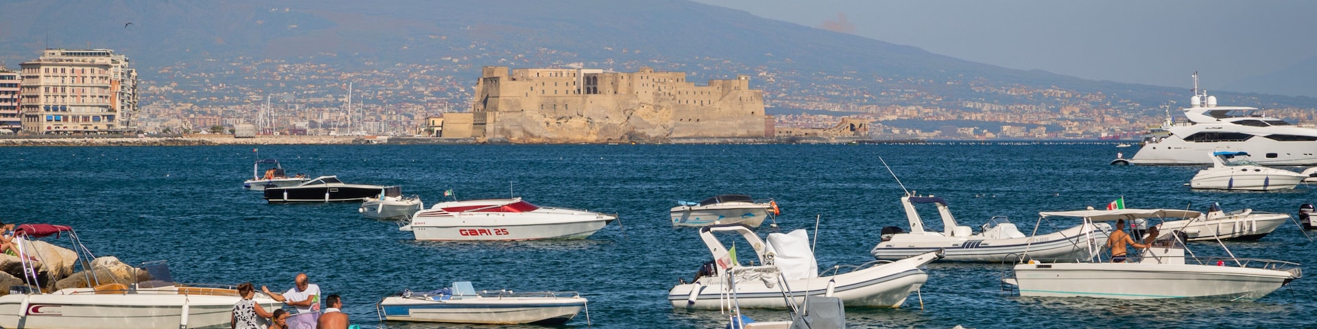 Uma marina com vários barcos, um castelo imponente e uma montanha ao fundo.