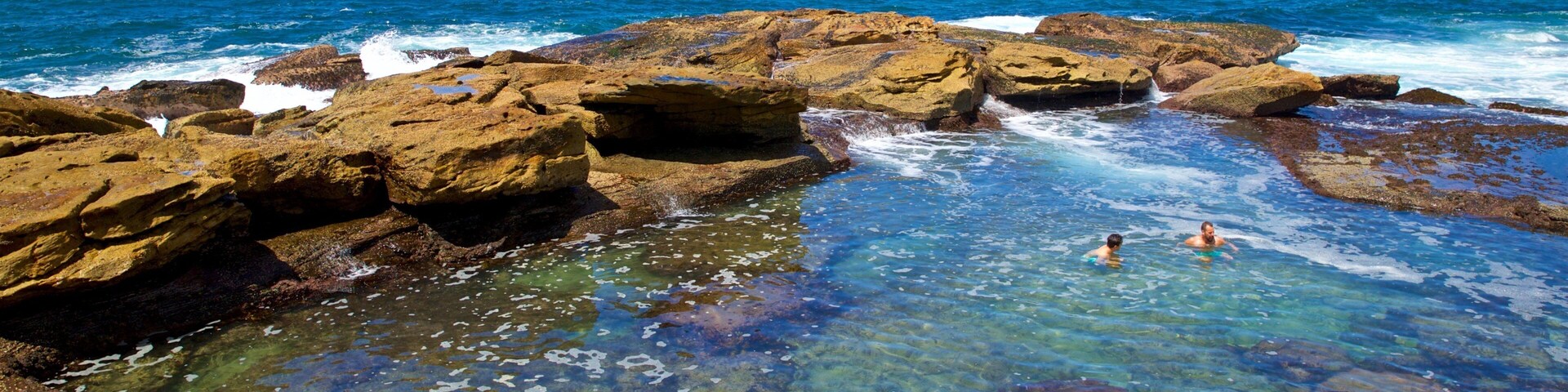 Litoral rochoso com águas cristalinas e duas pessoas a nadar.
