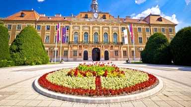Um edifício histórico com uma cúpula central, rodeado por um canteiro circular de flores vermelhas e brancas, e dois mastro com bandeiras.