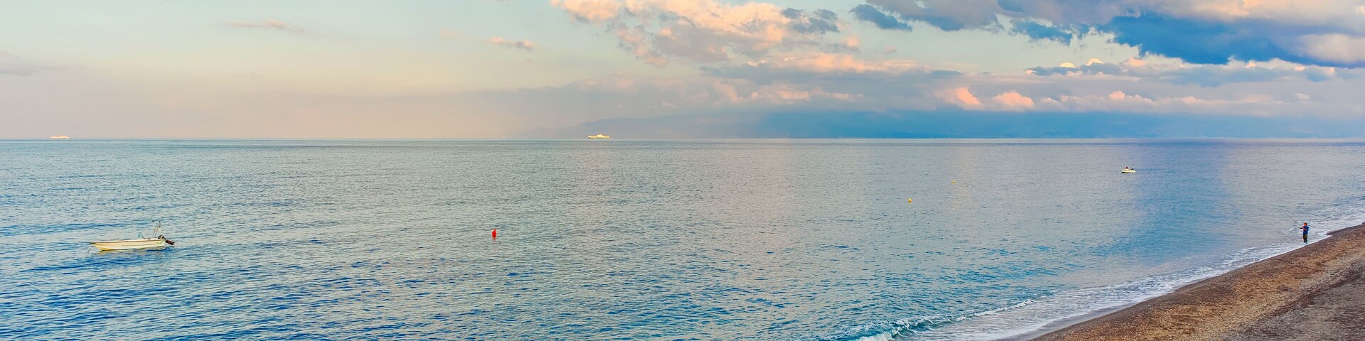 Uma praia com um barco na margem, um navio ao longe e uma pessoa pescando.