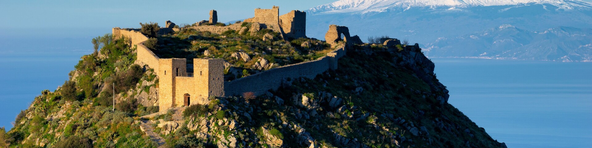 Um castelo de pedra histórico numa colina, com uma montanha nevada ao fundo.