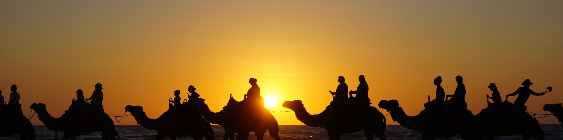 Um grupo de pessoas cavalgando camelos numa praia ao pôr do sol.