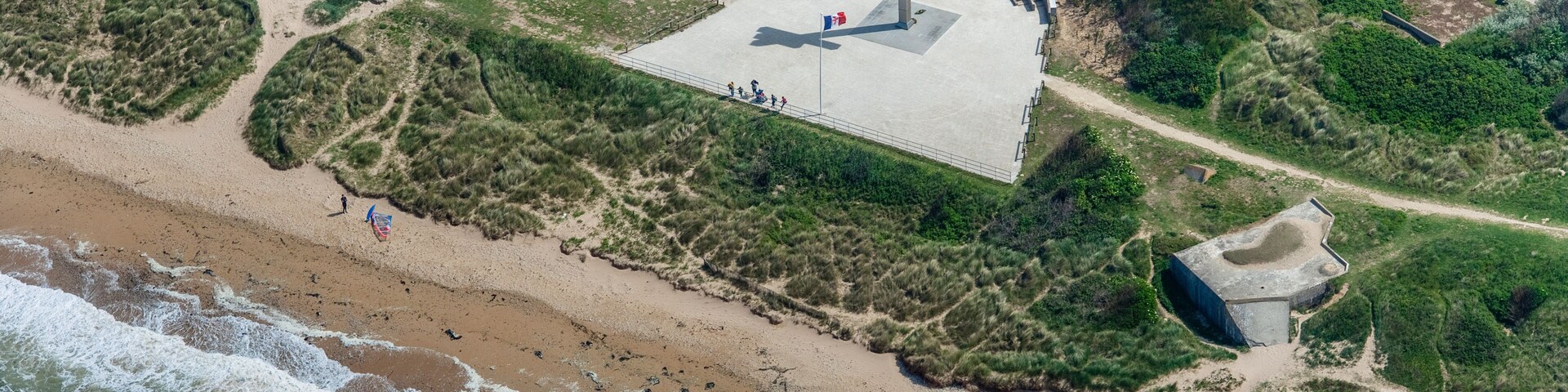 Vista aérea de uma área costeira com praia, um monumento comemorativo e um estacionamento.