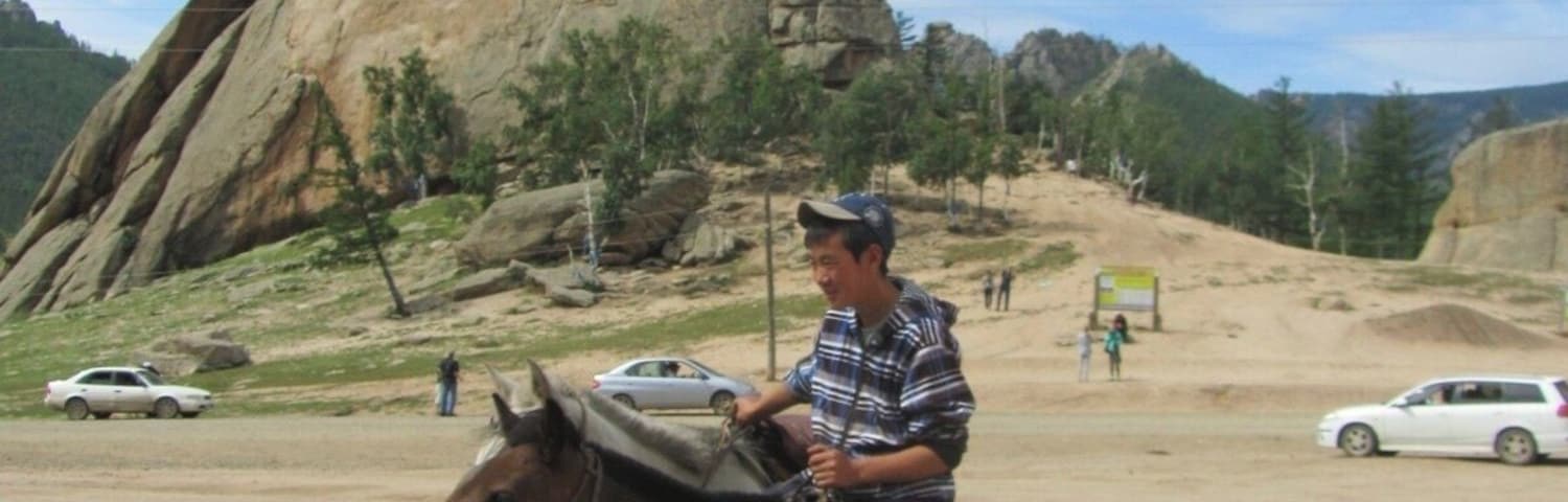 Uma pessoa a cavalo numa área arenosa, com uma grande formação rochosa ao fundo.